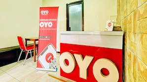 Uma área de recepção com balcão da marca OYO em vermelho e branco, um banner promocional, uma cadeira vermelha e uma parede revestida de azulejos.