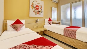 Quarto de hotel com duas camas, cada uma com roupa de cama branca e detalhes vermelhos, cabeceira de madeira, ar condicionado fixado na parede e janela com vidro jateado.