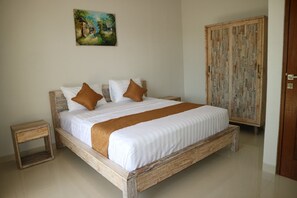 Uma cama de madeira com roupa de cama branca e almofadas castanhas, uma pequena mesa de cabeceira de madeira e um guarda-roupa de madeira.
