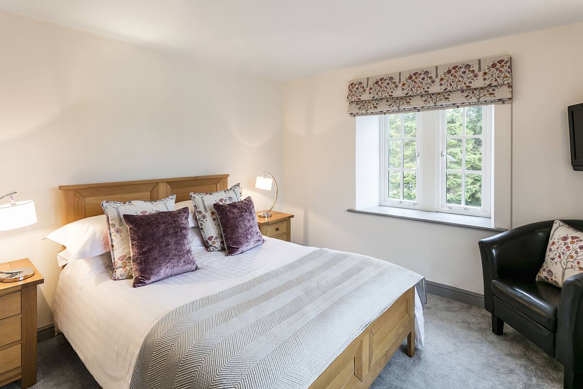 Quarto com cama de madeira, duas mesas de cabeceira, um criado-mudo, janela com valance floral e uma poltrona de couro preto.