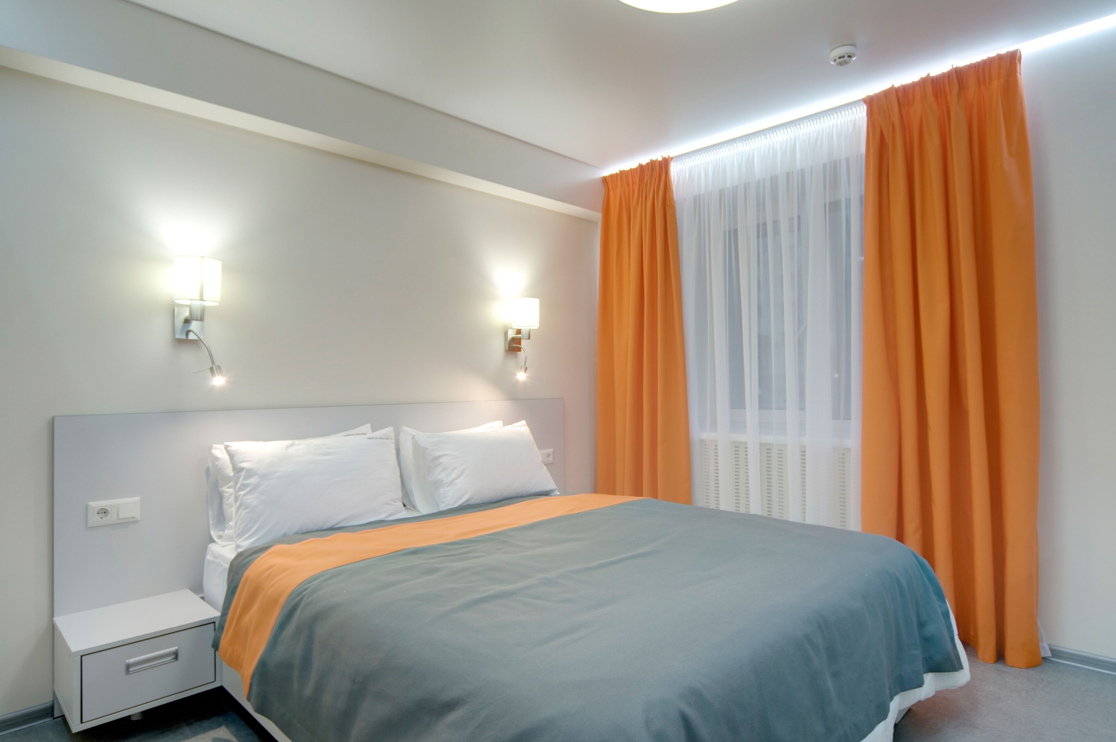 Quarto de hotel com cama, mesa de cabeceira, duas luminárias fixadas na parede e janela com cortinas laranja.