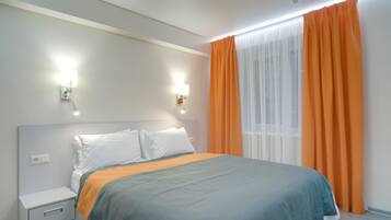 Quarto de hotel com cama, mesa de cabeceira, duas luminárias fixadas na parede e janela com cortinas laranja.
