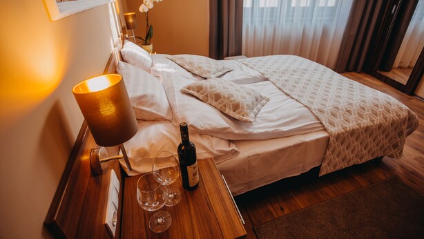 Cama bem arrumada com uma garrafa e copos em uma mesa de cabeceira, uma luminária e um quadro na parede.