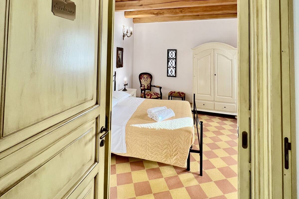 Quarto com piso xadrez, cama com colcha bege, teto de madeira e armário branco.