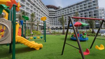 Um parquinho com baloiços e escorregas, rodeado por um hotel com várias varandas e uma piscina.