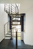 Escadaria em espiral com corrimãos de metal e proteção de vidro, que dá acesso a uma área externa com vista para edifícios.
