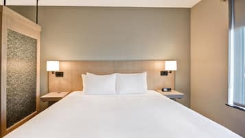 Quarto de hotel com uma cama grande, duas luminárias de cabeceira, uma mesinha de cabeceira e uma janela com persianas.