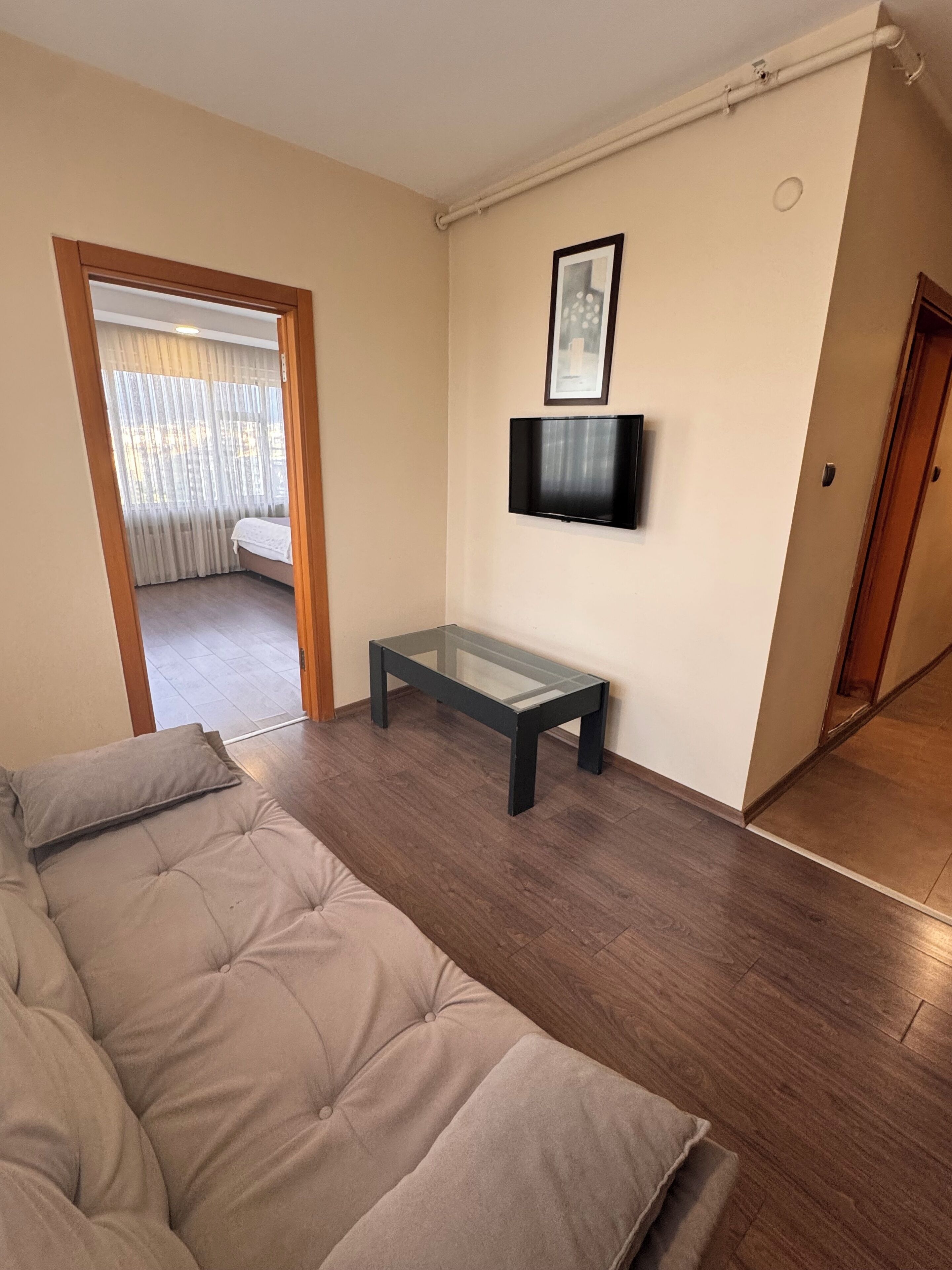 Sala de estar com um sofá bege, uma mesa de centro de vidro e uma televisão de tela plana na parede.