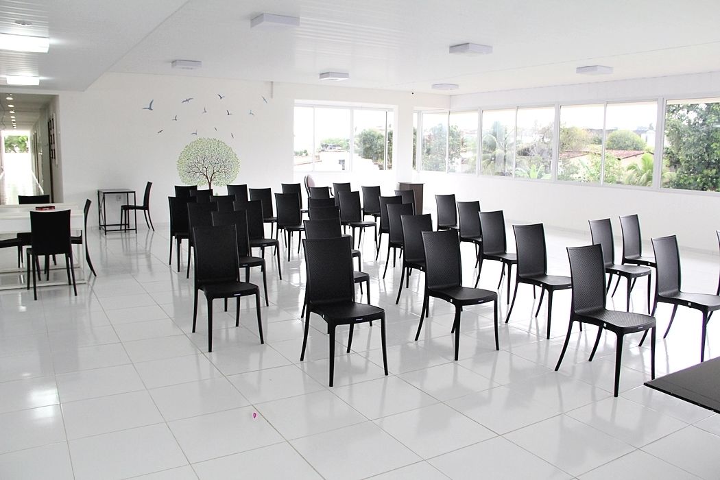 Uma sala de conferências com fileiras de cadeiras pretas e mesas, grandes janelas com vista para o verde, e um mural na parede com pássaros voando ao redor de uma árvore.