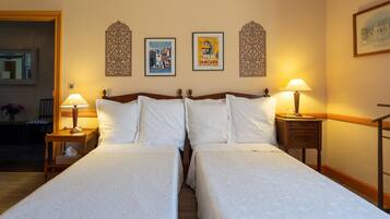 Uma cama de casal com roupa de cama branca, duas mesas de cabeceira com abajures e três quadros na parede.