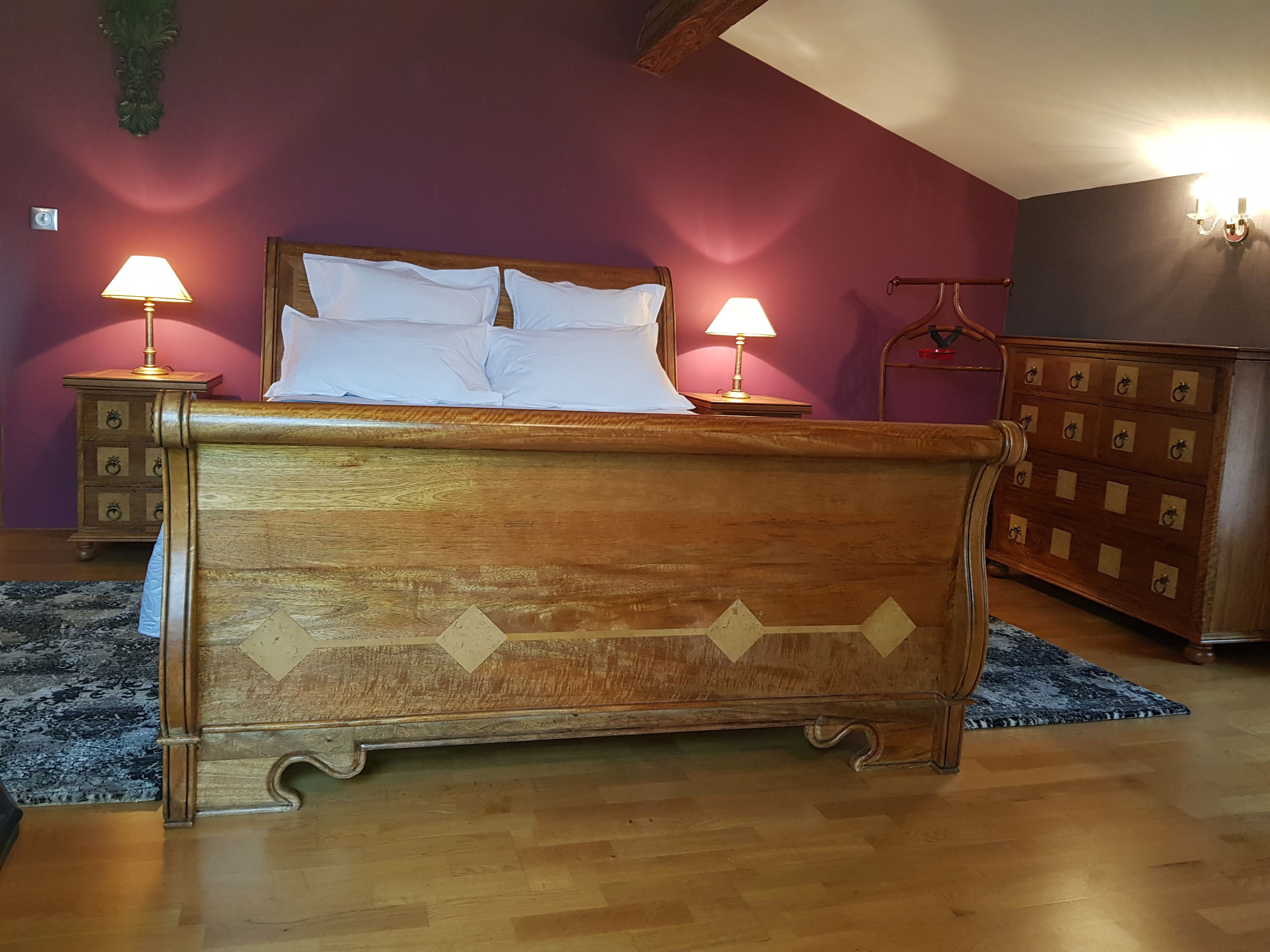 Uma cama de madeira com cabeceira, duas luminárias de apoio, um penteado e um tapete estampado em um quarto com paredes roxas.