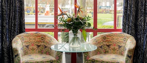 Quarto com duas poltronas estampadas de flores viradas para uma mesa de vidro com um vaso de flores. Janelas amplas com molduras vermelhas oferecem vista para o exterior.