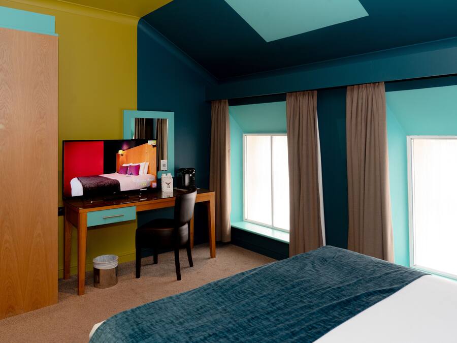 Quarto de hotel com uma cama, uma escrivaninha com cadeira, uma pequena lixeira e uma janela com cortinas.