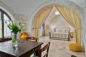 Um interior rústico com arcos de pedra, uma mesa de jantar de madeira e uma cama com cabeceira branca.