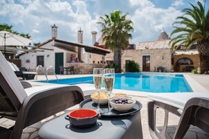 Mesa à beira da piscina com bandeja de comida e bebidas, com vista para uma piscina e um edifício de pedra ao fundo.