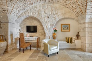 Quarto de paredes de pedra com televisão, uma cadeira de vime, um sofá e uma mesa com um vaso de flores.