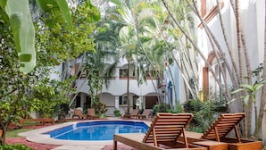 Uma área à beira da piscina com espreguiçadeiras, piscina e um edifício com arcos ao fundo.