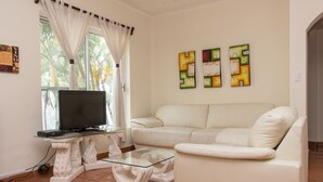 Uma sala de estar com televisão sobre uma mesa de vidro, um sofá branco e três quadros abstratos na parede.