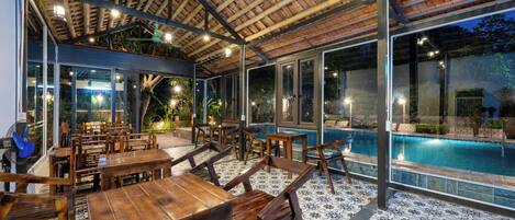 Uma área de refeições ao ar livre com mesas e cadeiras de madeira, piscina e teto coberto.