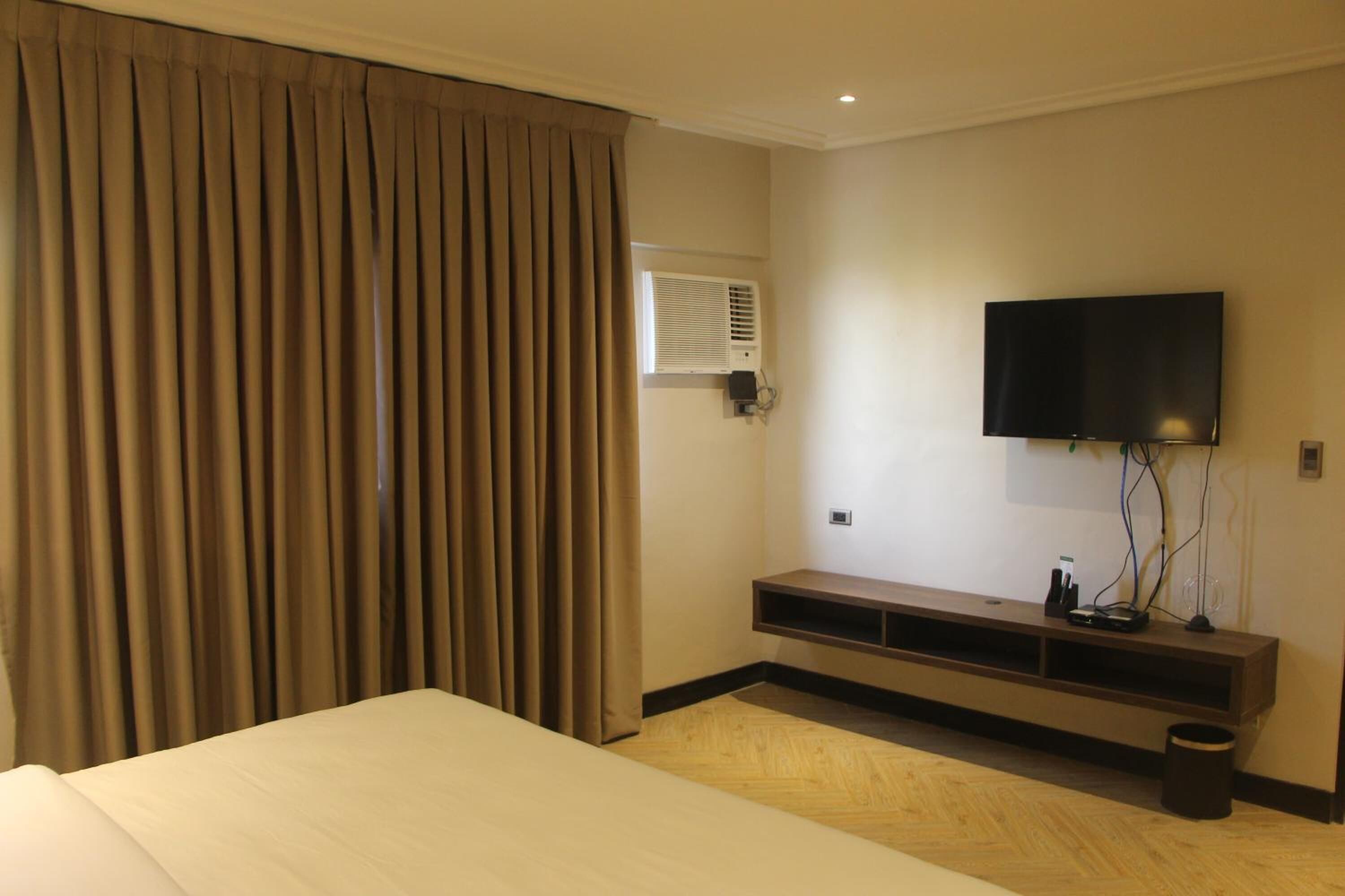 Quarto de hotel com cama, televisão na parede, pequena mesinha de apoio e janela com cortinas.