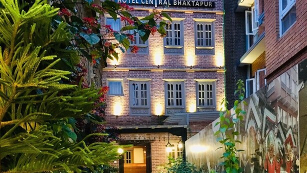 Entrada de hotel com fachada de tijolos, uma placa com a inscrição 'Hotel Central Ral Bhaktapur' e um caminho ladeado por vegetação.