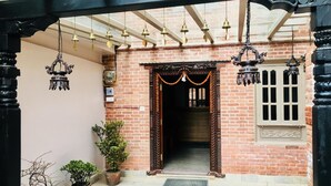 Uma entrada tradicional com parede de tijolos, porta de madeira e portão de metal preto. Há sinos pendurados e uma pequena luminária decorativa.