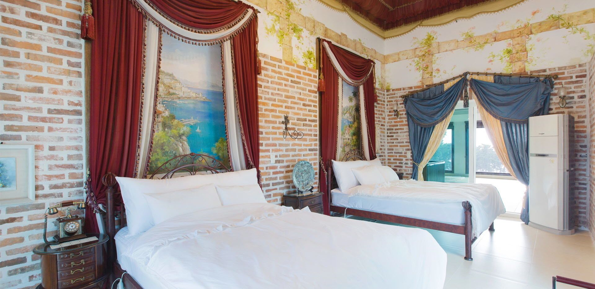 Quarto de hotel com duas camas, parede de tijolos, um mural e uma janela com cortinas.