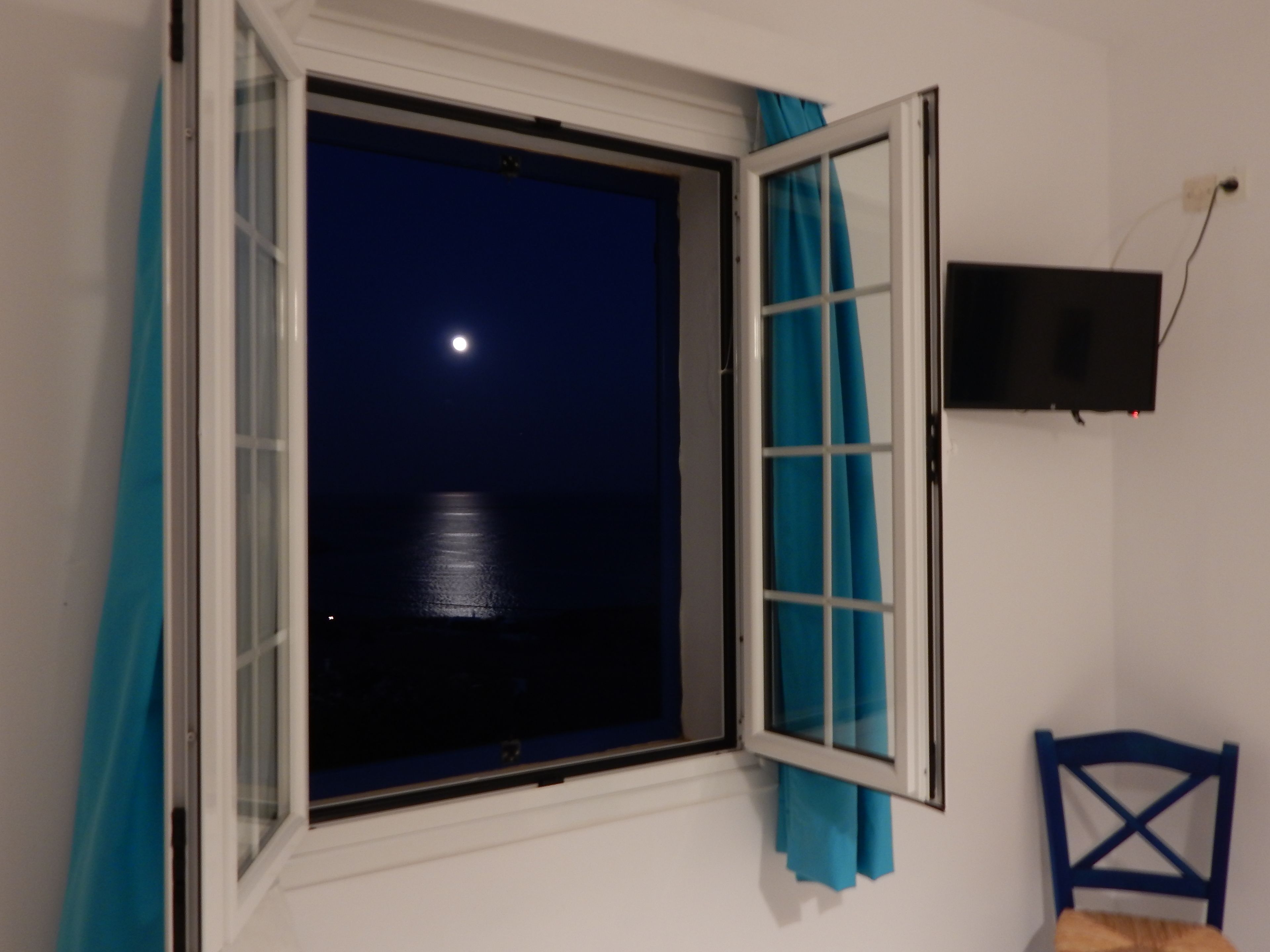 Quarto com janela aberta para o exterior, mostrando um céu noturno com lua e vista para o mar.