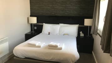 Cama bem arrumada com lençóis brancos e duas toalhas, cabeceira escura, duas mesas de cabeceira com luminárias e uma janela com cortinas.
