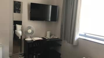 Quarto de hotel com cama, escrivaninha com cadeira, televisão, ventilador e janela com cortinas.