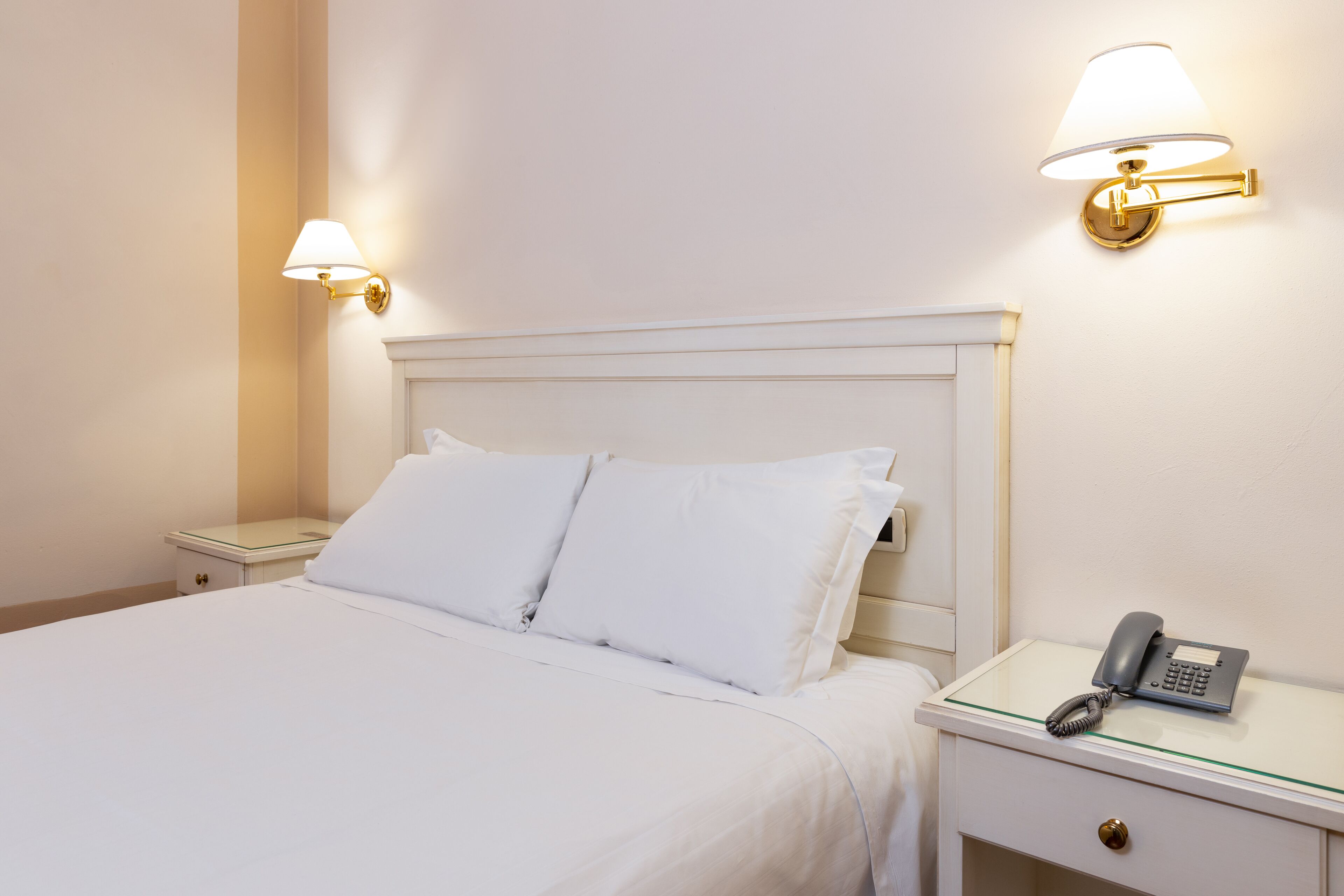Quarto de hotel com cama bem arrumada, telefone de apoio e duas luminárias fixadas na parede.