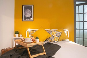 Quarto bem arrumado com uma cama, uma mesinha de cabeceira com bandeja, um quadro na parede e uma janela com vista.
