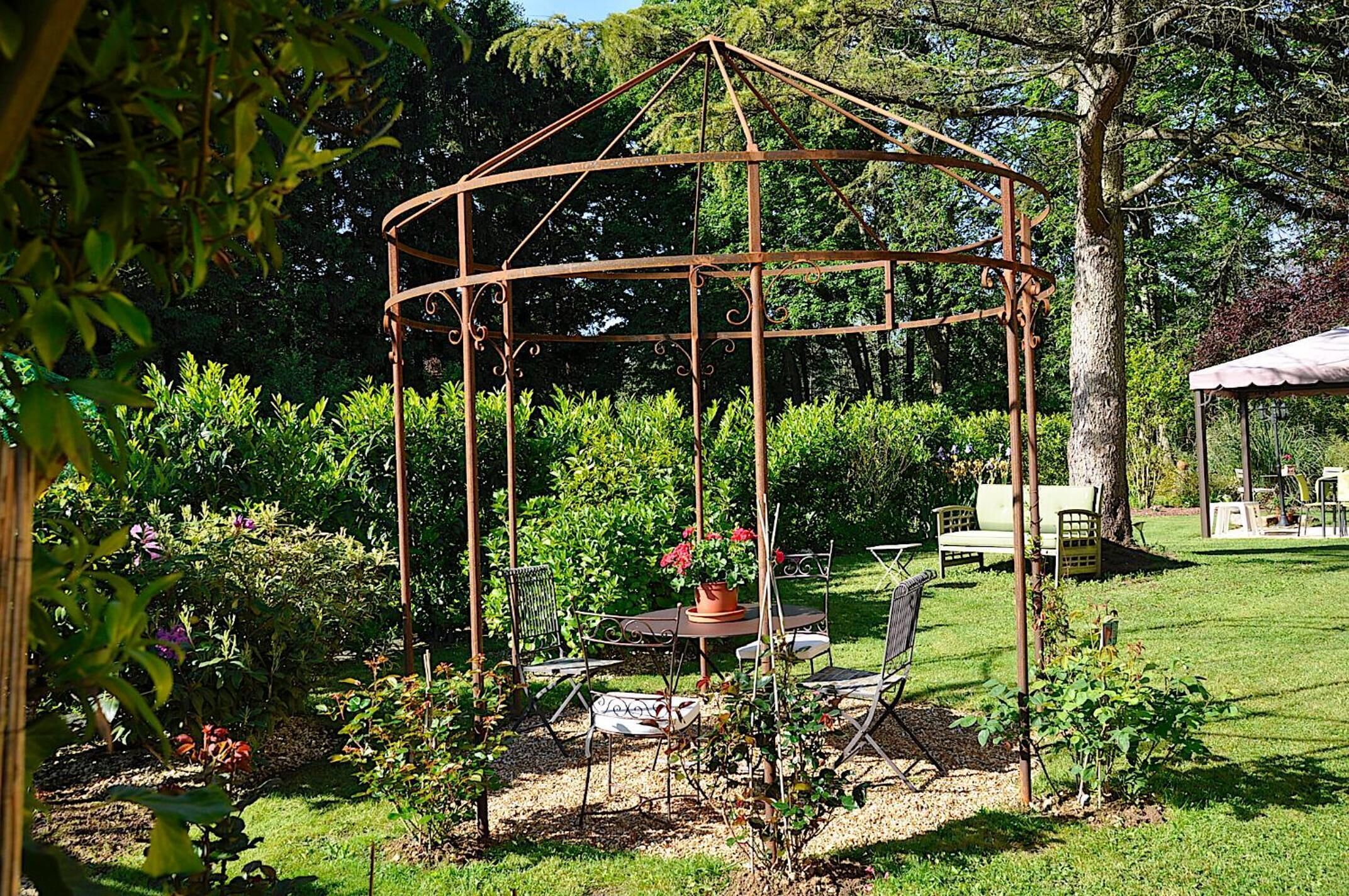 Um gazebo com estrutura metálica, rodeado por um jardim com diversas plantas e uma mesa com cadeiras.