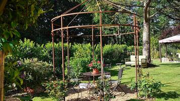 Um gazebo com estrutura metálica, rodeado por um jardim com diversas plantas e uma mesa com cadeiras.
