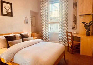 Quarto com cama, mesa de cabeceira, abajur, cadeira, escrivaninha, planta em vaso e janela com cortinas.
