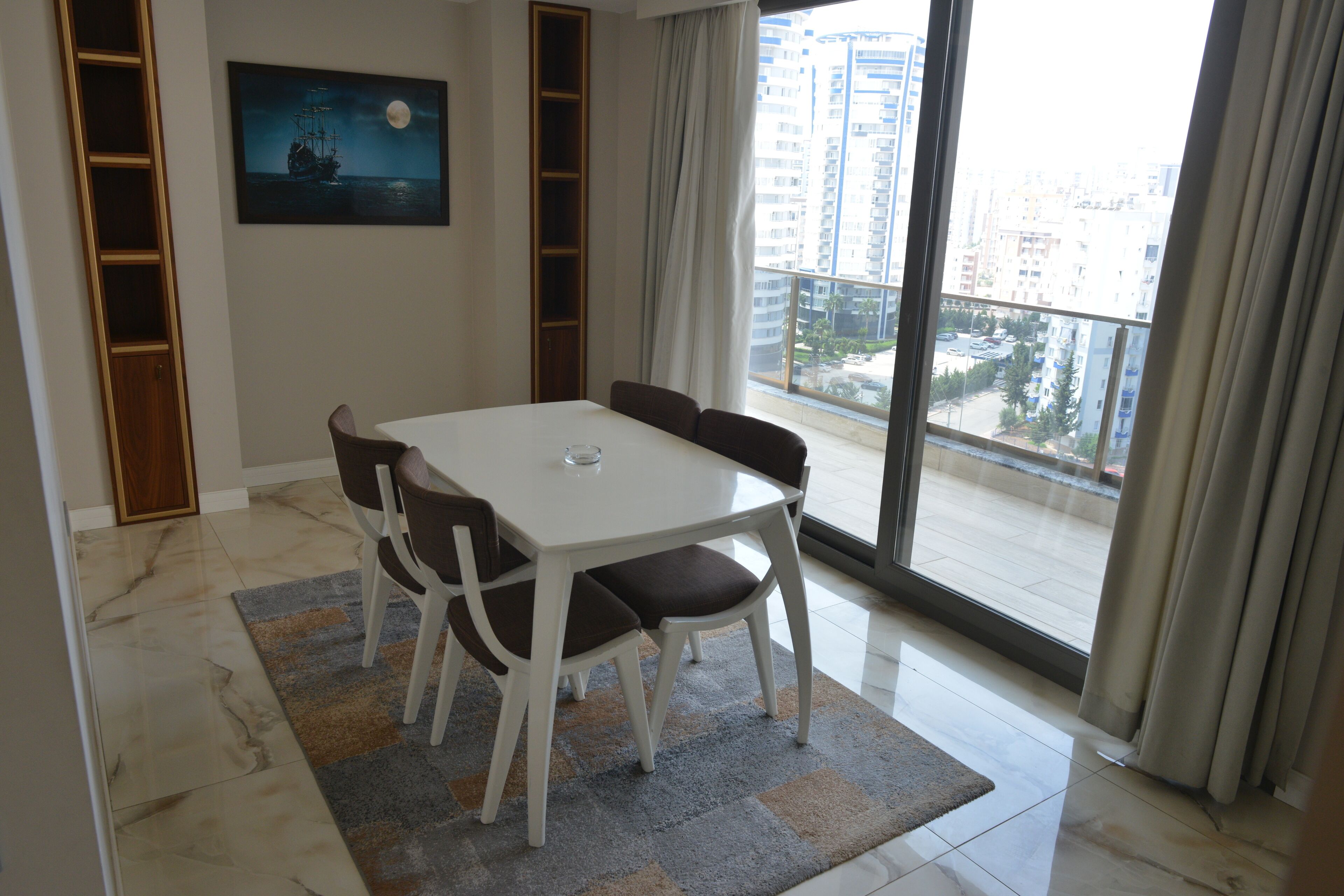 Uma sala de jantar com uma mesa e cadeiras brancas, um quadro na parede e vista para edifícios através de grandes janelas.