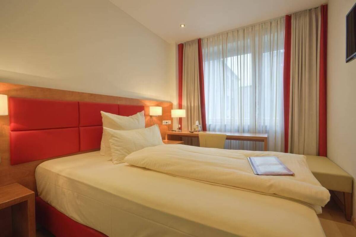 Quarto de hotel com uma cama grande, mesas de cabeceira, uma escrivaninha e uma janela com cortinas.