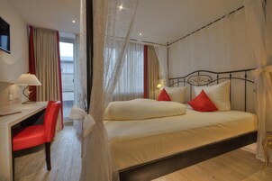 Quarto de hotel com cama, escrivaninha, cadeira e janela com cortinas.