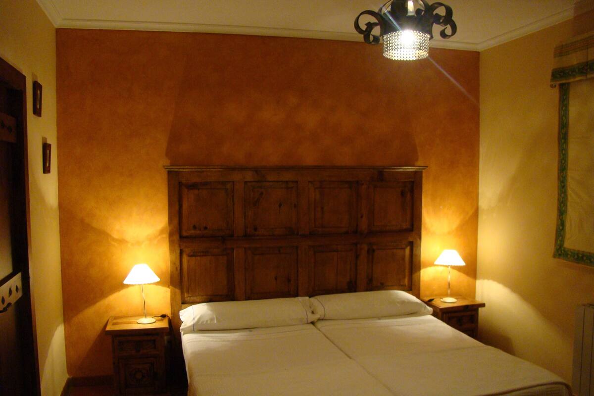 Quarto com cama de casal, duas luminárias laterais, cabeceira de madeira e luminária no teto.