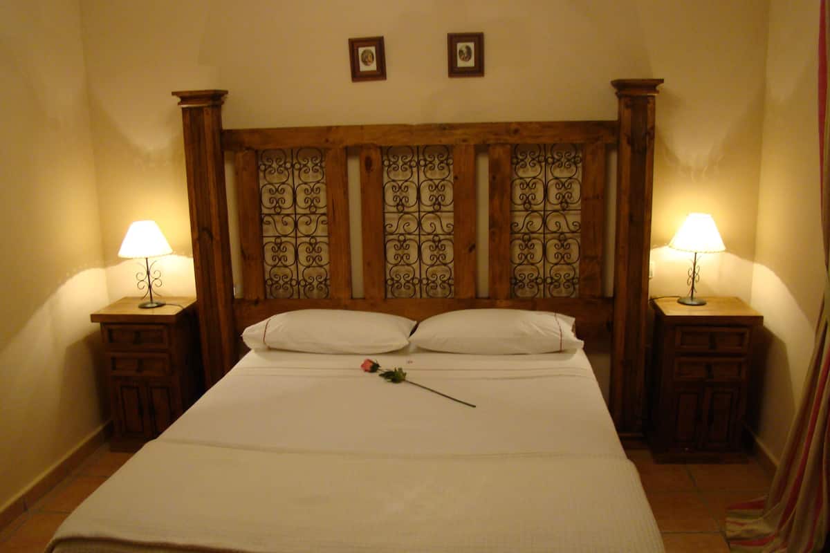 Cama bem arrumada com uma única rosa, duas luminárias de cabeceira e cabeceira de madeira com detalhes metálicos elaborados.