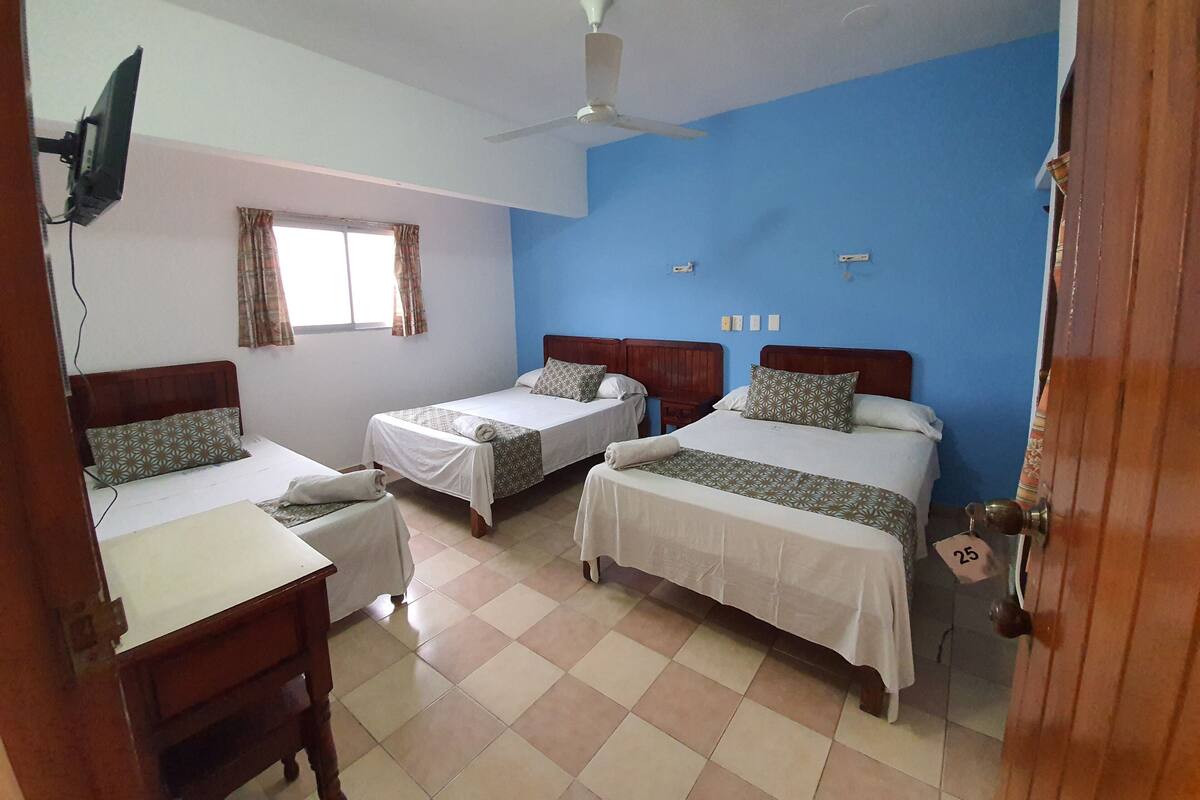 Quarto de hotel com duas camas de solteiro, uma escrivaninha de madeira, um ventilador de teto, uma janela com cortinas e piso de azulejos.