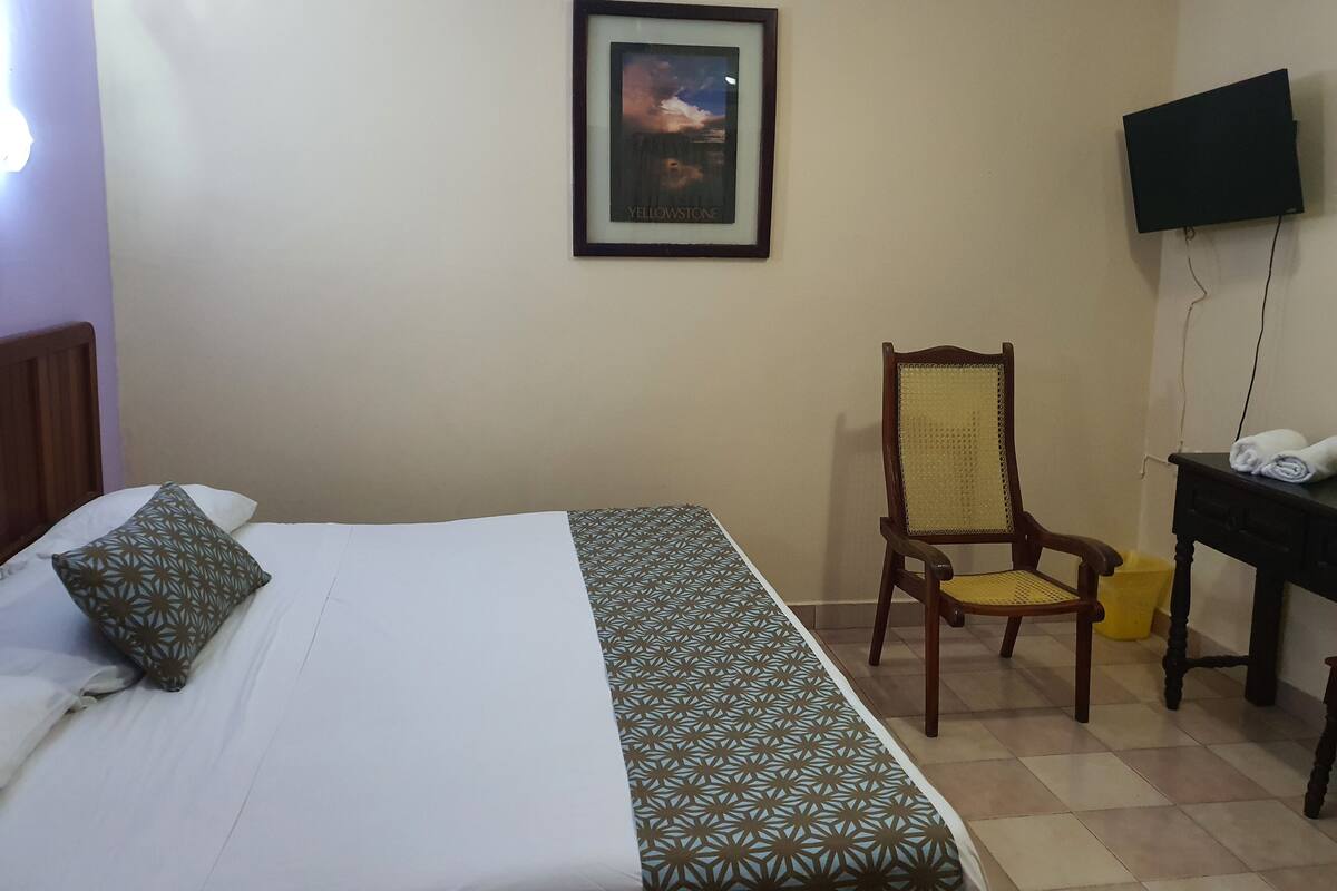 Quarto de hotel com cama, poltrona, escrivaninha, televisão e um quadro pendurado na parede.