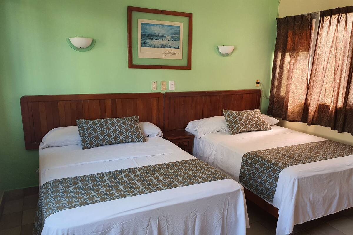 Quarto de hotel com duas camas, cada uma com colcha e almofadas estampadas, cabeceira de madeira, um quadro pendurado na parede e uma janela com cortinas.