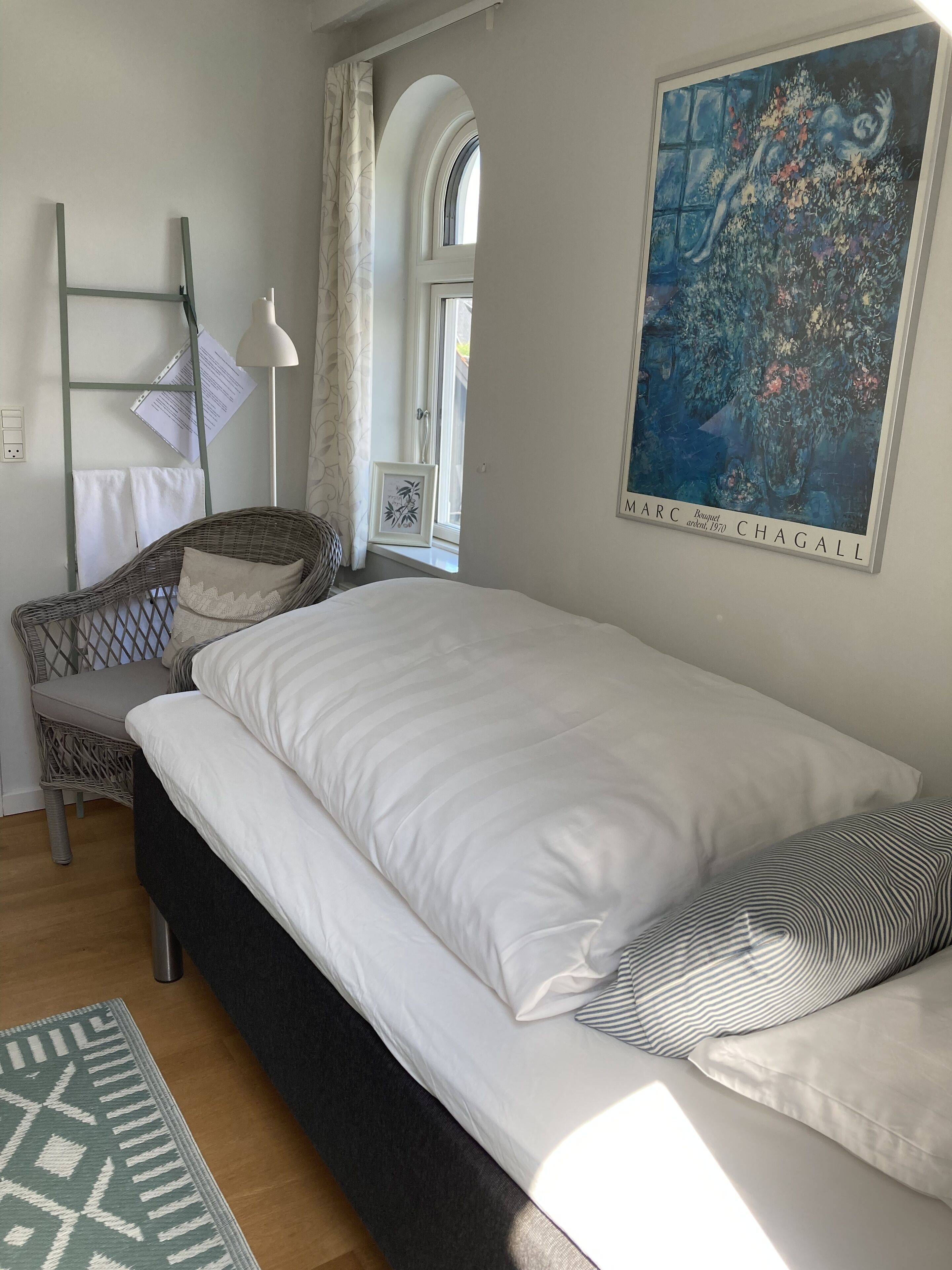 Um quarto com cama, uma cadeira de vime, um abajur e um quadro na parede.