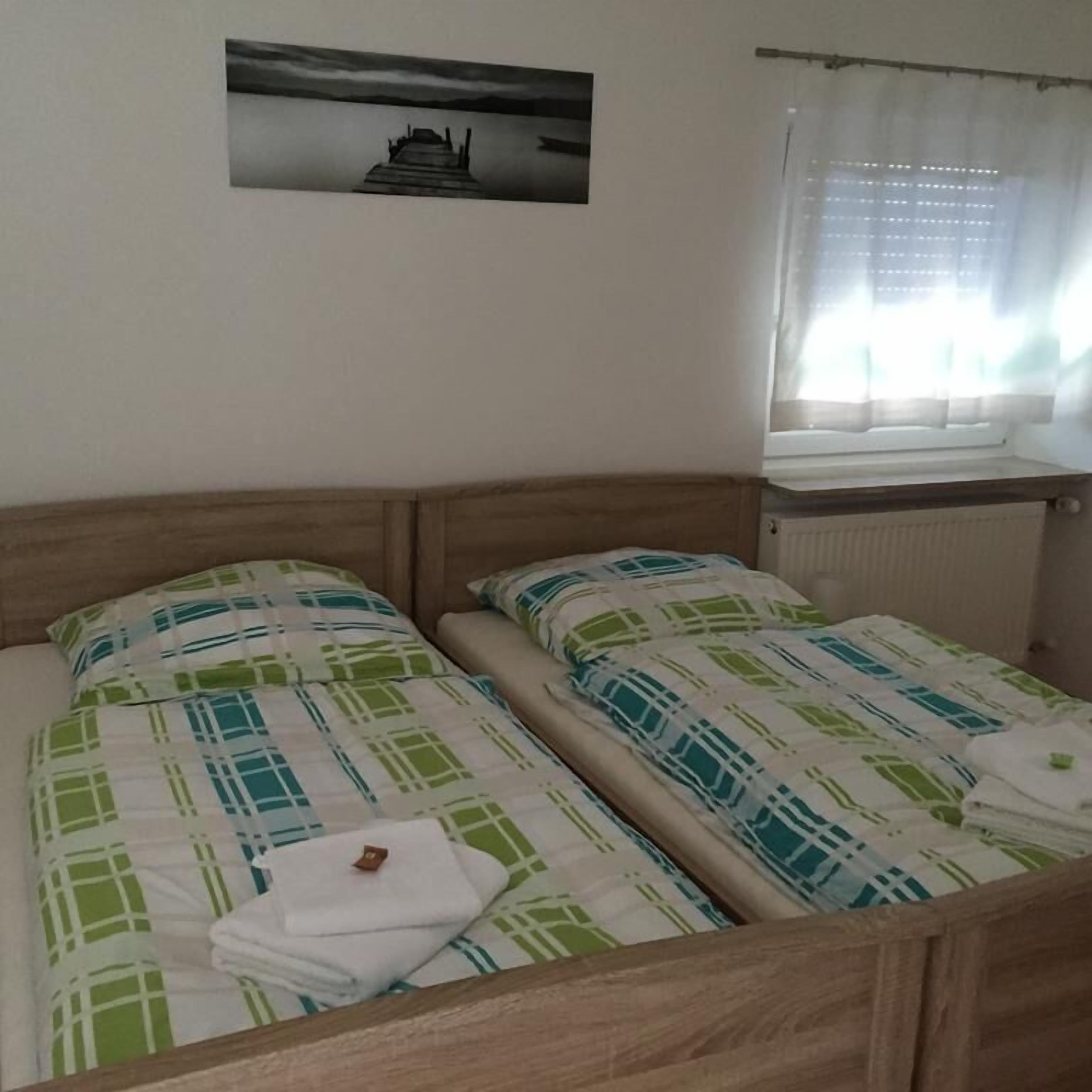 Quarto com duas camas de solteiro, cada uma com roupa de cama e almofadas estampadas, cabeceira de madeira, janela com cortinas leves e uma moldura com imagem na parede.