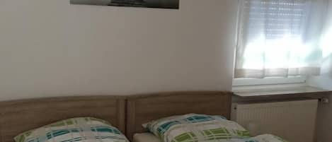 Quarto com duas camas de solteiro, cada uma com roupa de cama e almofadas estampadas, cabeceira de madeira, janela com cortinas leves e uma moldura com imagem na parede.
