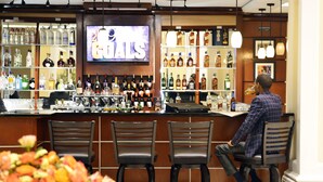 Uma área de bar com uma tela de TV exibindo 'GOALS' e prateleiras cheias de diversas garrafas de bebidas.