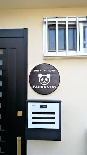 Uma porta preta com uma janela acima, uma placa com a inscrição "Town Cottage Panda Stay" e uma abertura para correspondência com a etiqueta "POST".