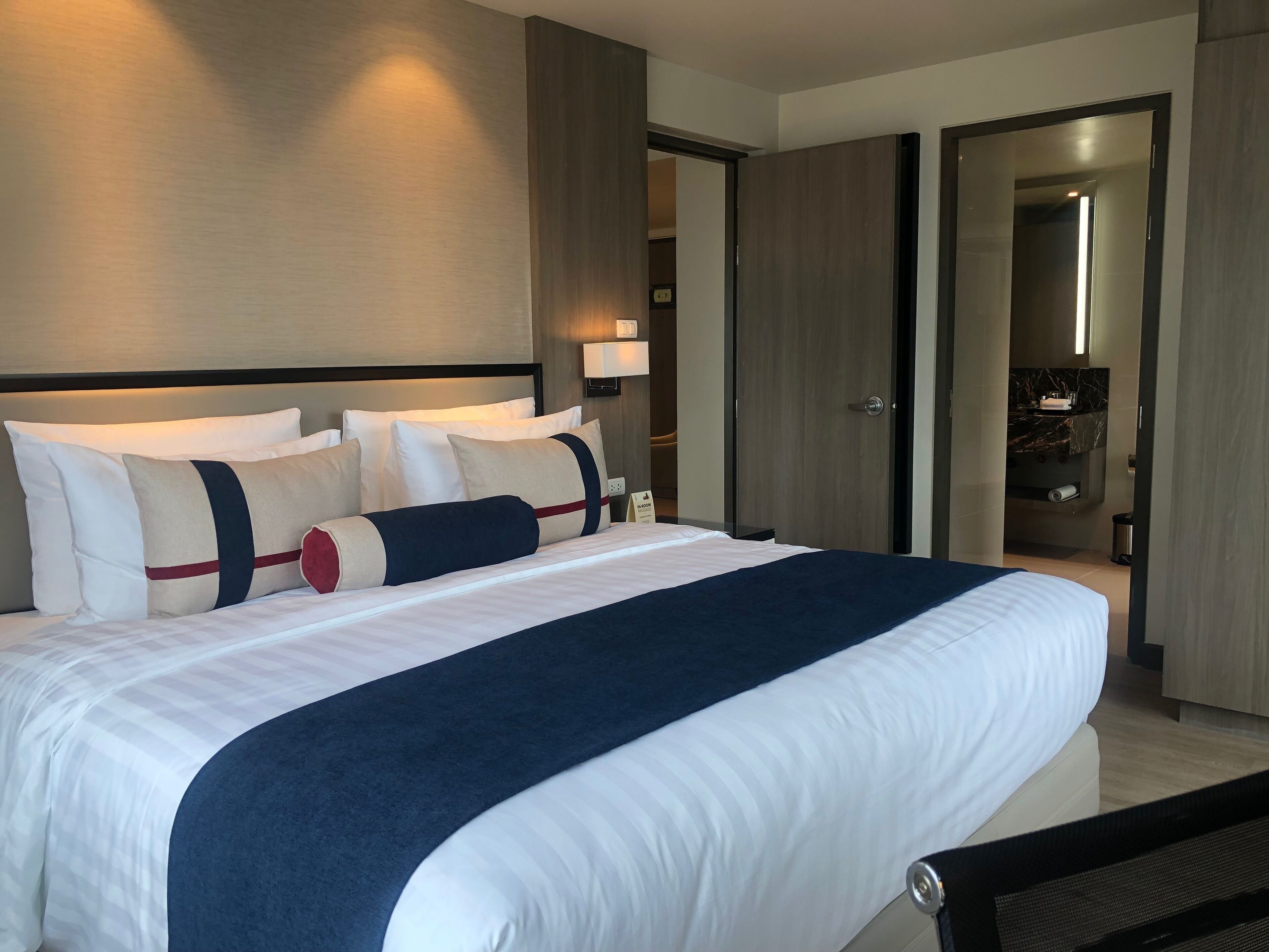 Quarto de hotel com uma cama grande, cabeceira, mesa de cabeceira, luminária e porta para um banheiro.