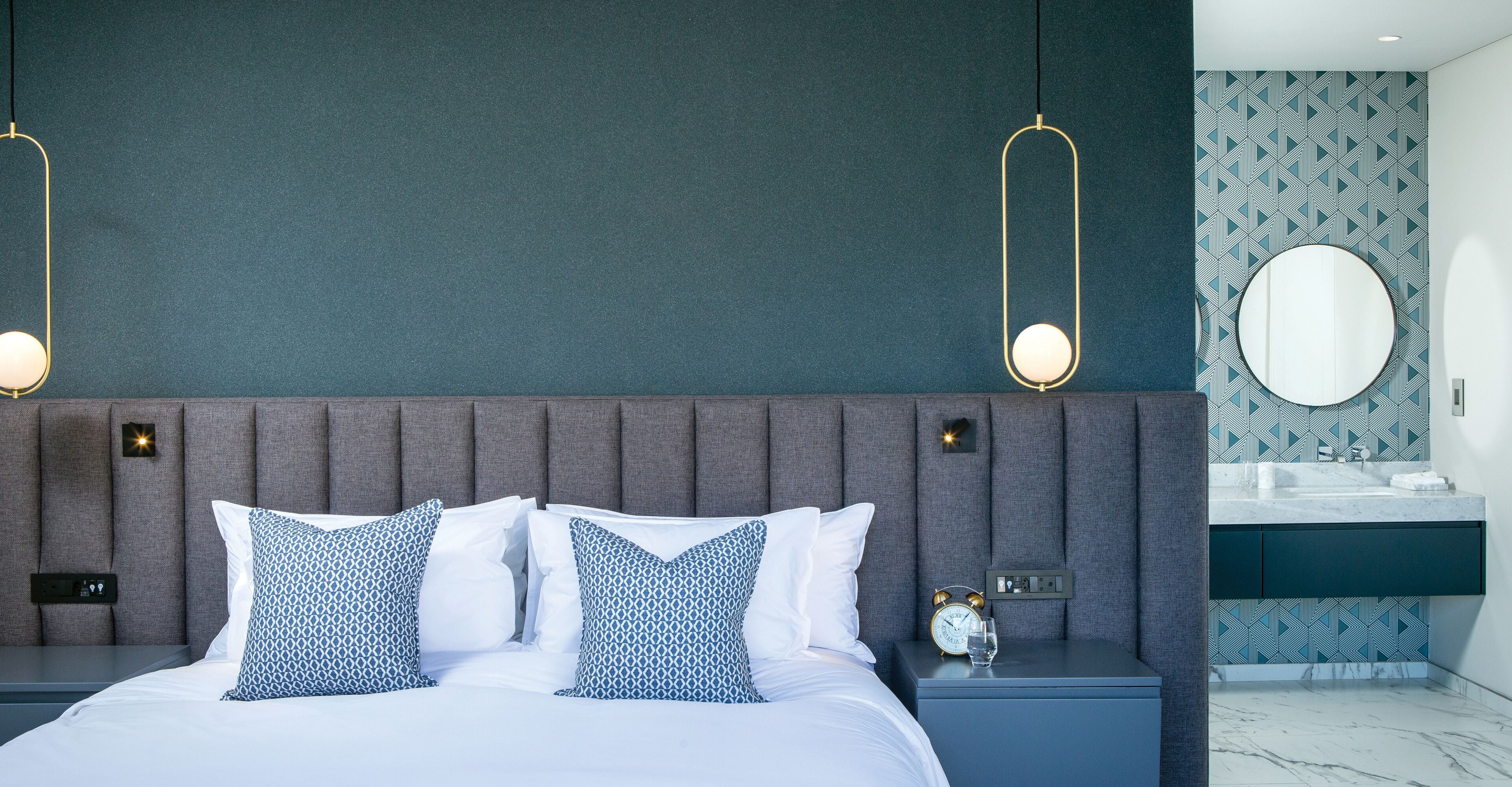 Quarto de hotel moderno com uma cama grande, duas mesas de cabeceira, um criado-mudo com relógio e banheiro com espelho e pia.
